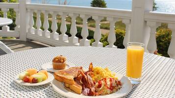 Uma mesa de café da manhã com torradas, ovos, bacon, frutas e um copo de suco de laranja em uma mesa branca com vista para o oceano.