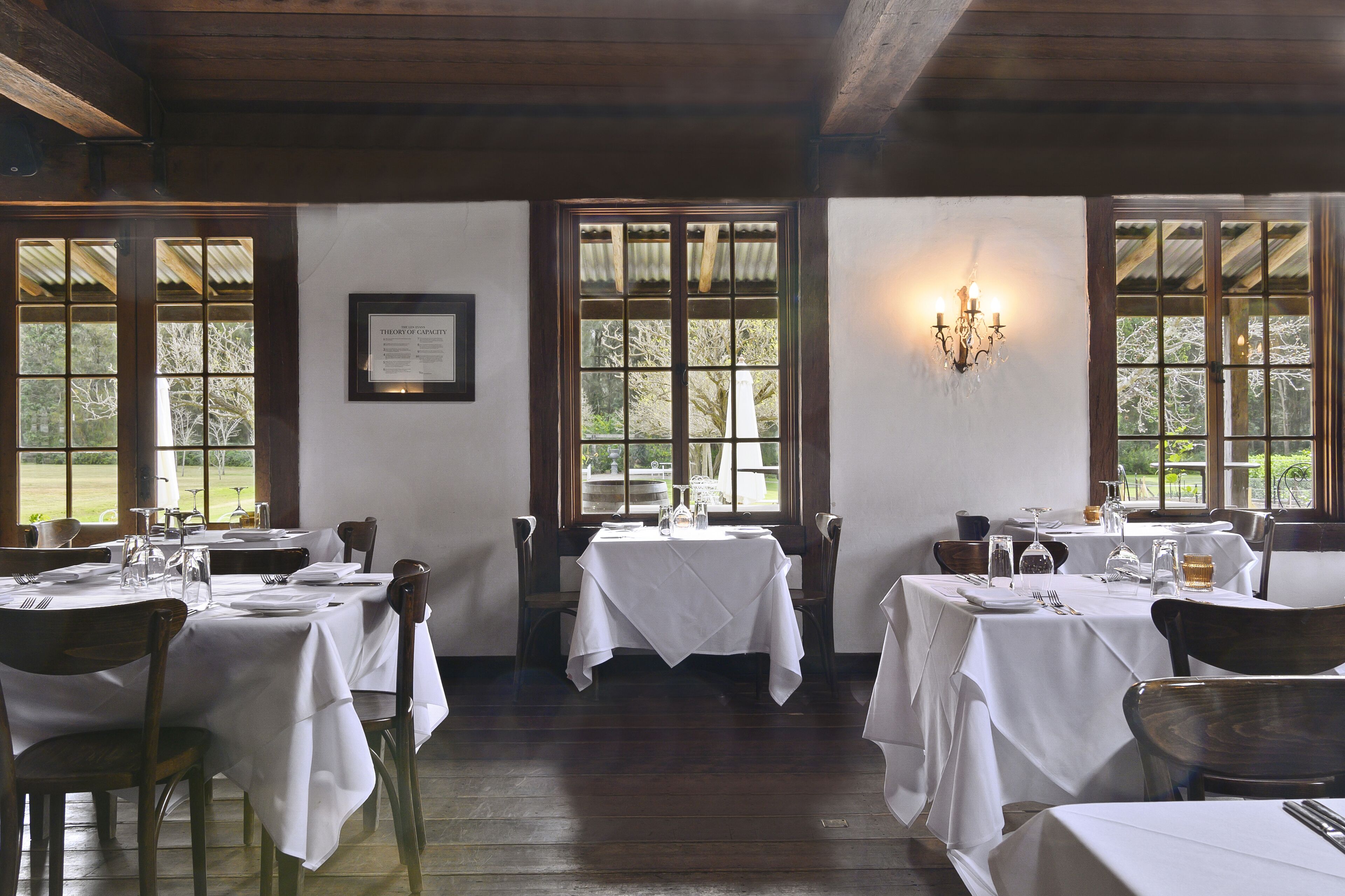 Uma sala de jantar com mesas postas para quatro pessoas, cadeiras de madeira e um lustre.