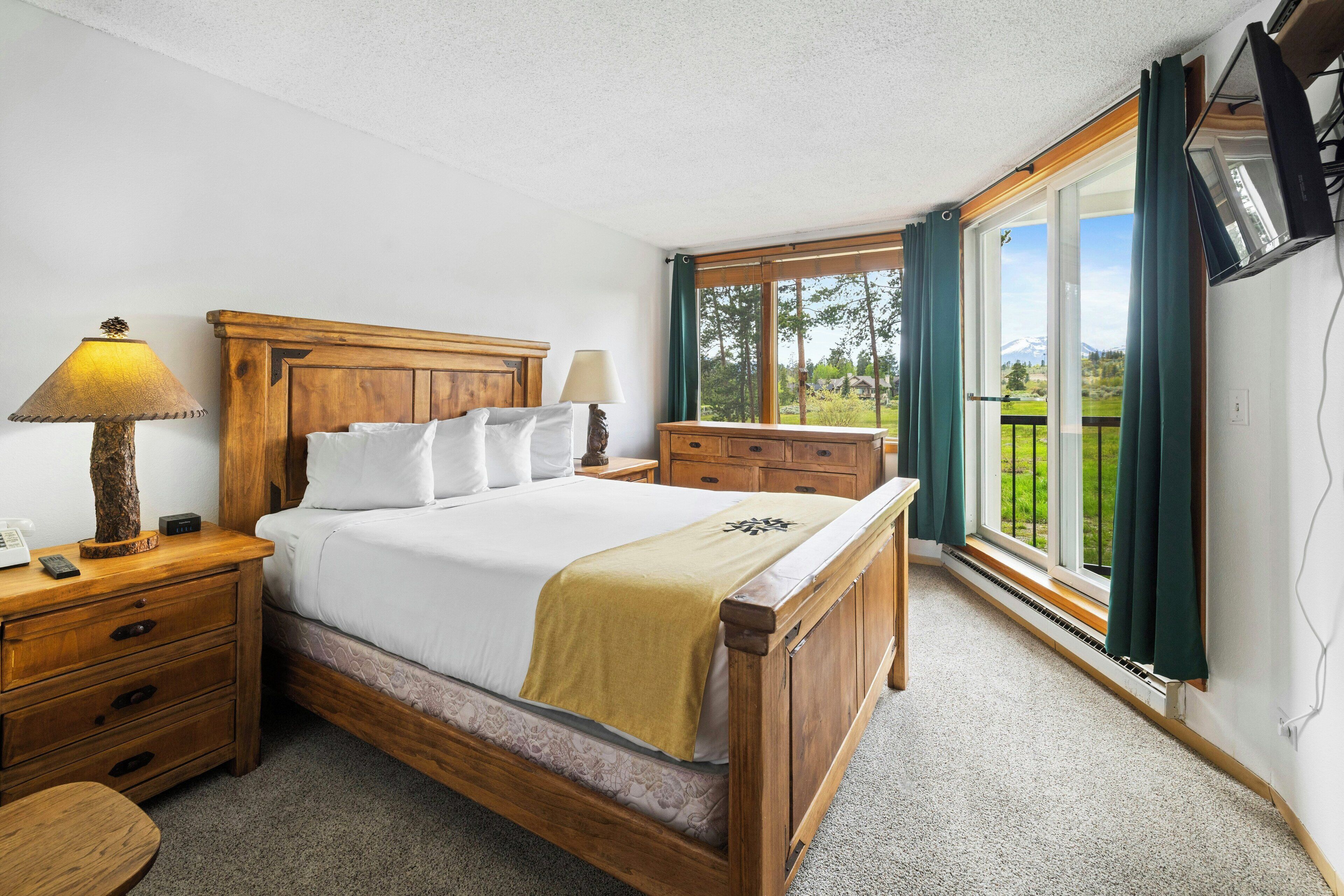 Uma cama de madeira com cabeceira, mesas de cabeceira, uma luminária e vista para uma varanda com cortinas.