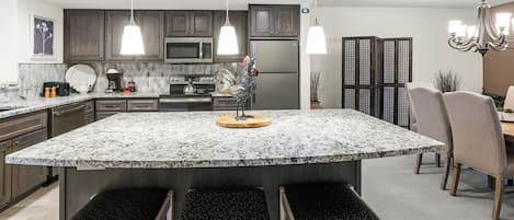 Uma cozinha com uma ilha grande, armários de madeira escura, eletrodomésticos de aço inoxidável, luminárias pendentes e um espaço de jantar com cadeiras e mesa.