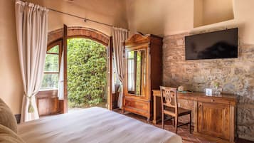Quarto com cama, escrivaninha e televisão. Cortinas nas janelas e parede de pedra à vista.