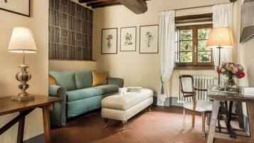 Quarto aconchegante com mesa de madeira, sofá azul, um banquinho bege e uma cadeira. Há gravuras botânicas emolduradas na parede, um radiador e uma janela com cortinas.