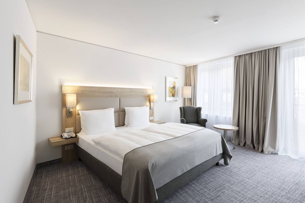 Quarto de hotel com uma cama grande, mesas de cabeceira, uma cadeira, uma mesinha redonda e uma janela ampla com cortinas.