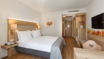 Quarto de hotel com cama, luminárias de cabeceira, escrivaninha com telefone, televisão e um quadro na parede.