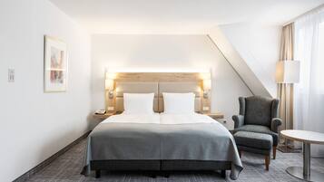 Quarto de hotel com uma cama grande, uma cadeira, uma mesinha e um abajur.