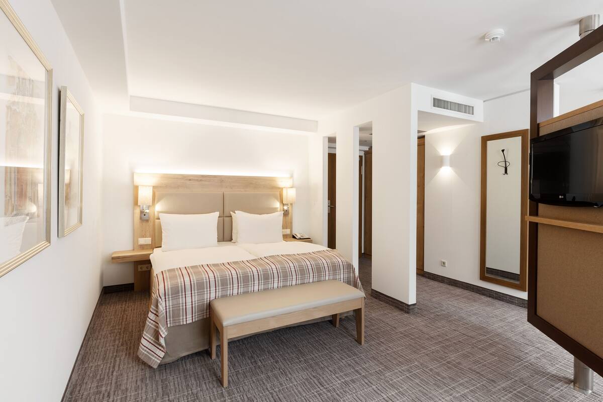 Quarto de hotel com uma cama grande, mesas de cabeceira, um banco, uma televisão e um espelho.