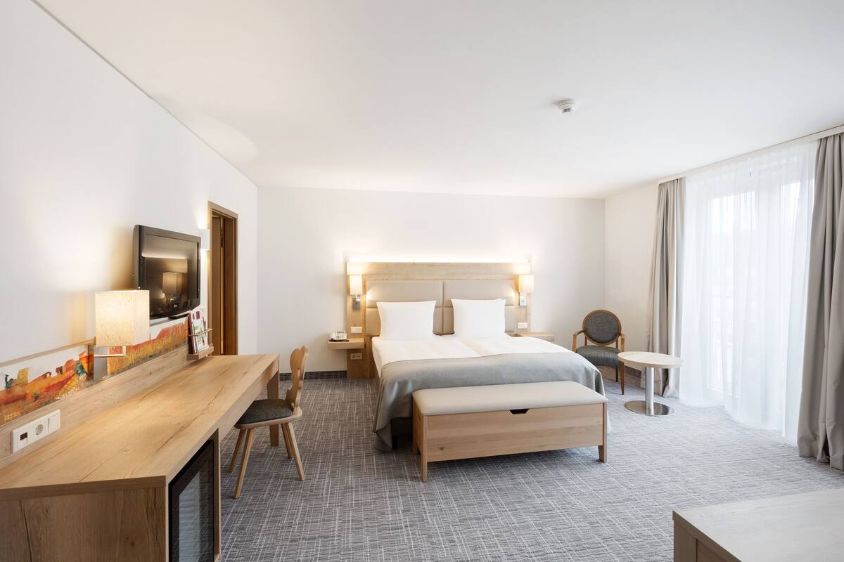Quarto de hotel com cama, mesas de cabeceira, escrivaninha, cadeira, televisão e janela com cortinas.