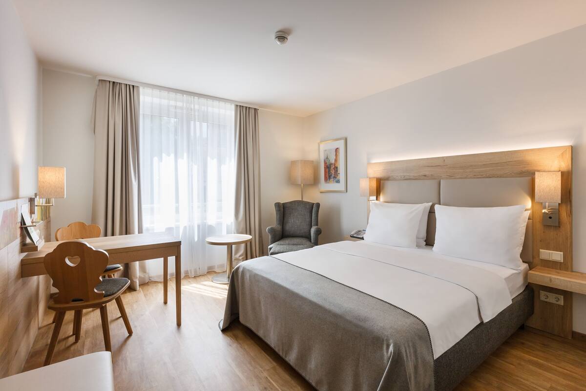 Quarto de hotel com cama, escrivaninha, cadeira, luminária e um quadro na parede.