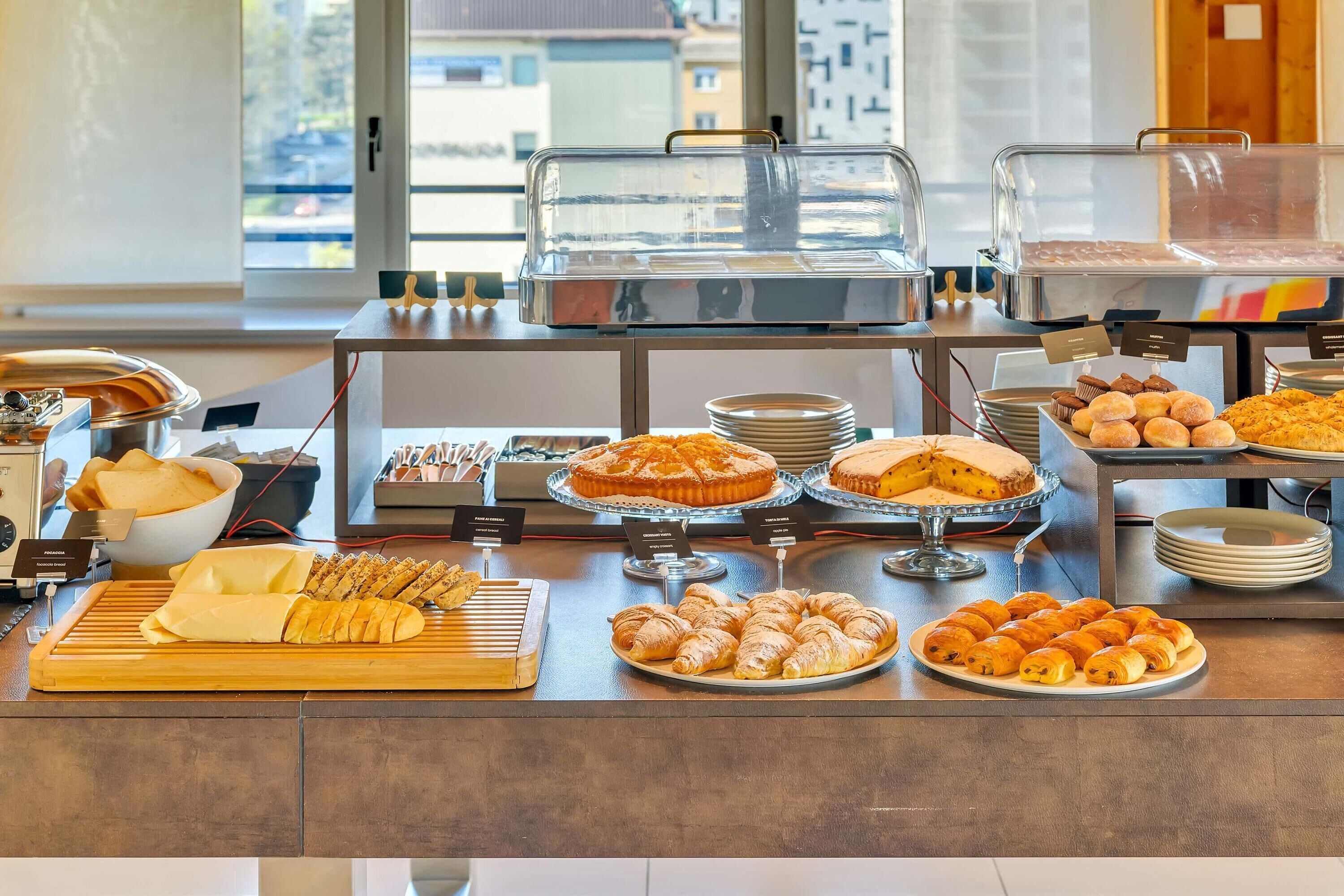 Buffet com diversos produtos de panificação, incluindo pastéis e tortas, disposto num balcão com vista para a cidade através de grandes janelas.