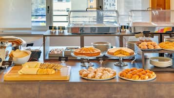 Buffet com diversos produtos de panificação, incluindo pastéis e tortas, disposto num balcão com vista para a cidade através de grandes janelas.
