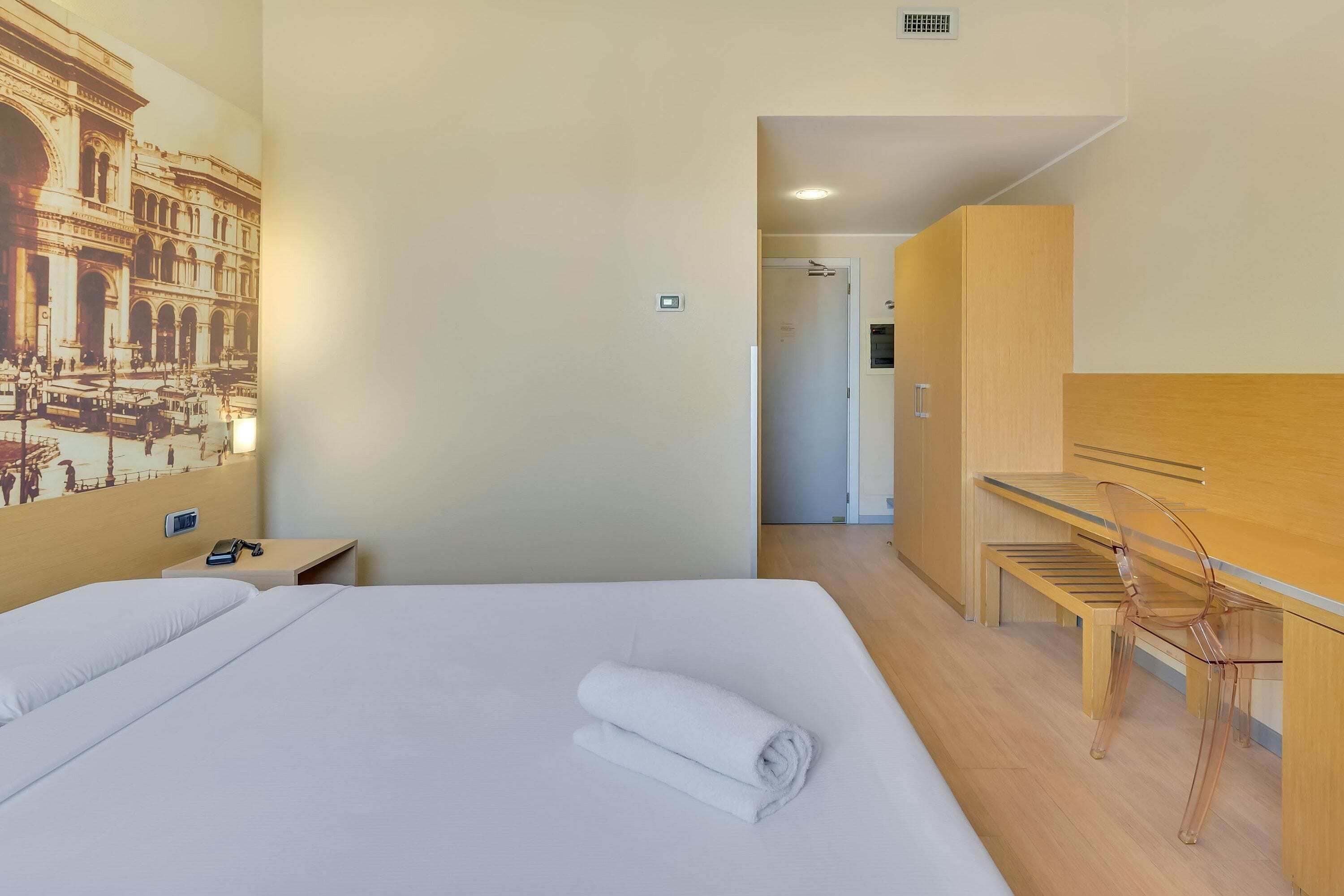 Quarto de hotel com cama, escrivaninha, cadeira e telefone fixado na parede.