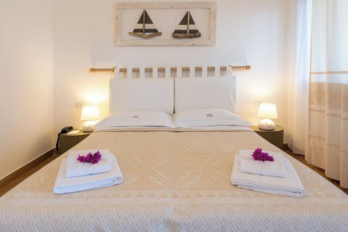 Uma cama de casal bem arrumada com um espalhador bege texturizado, dois travesseiros e duas toalhas dobradas com flores roxas em um quarto de hotel.