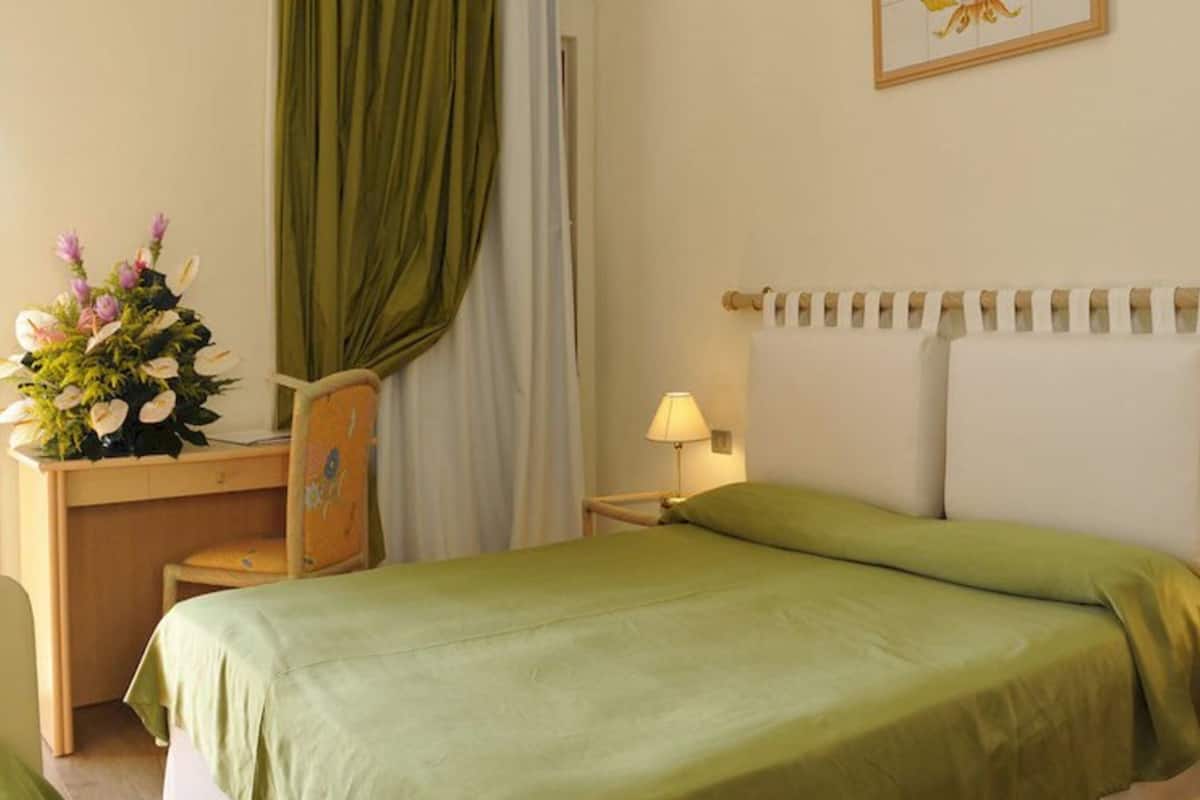 Quarto de hotel com uma cama, duas luminárias laterais, uma mesa de cabeceira com abajur, uma cadeira, uma escrivaninha com um arranjo floral e uma cortina.