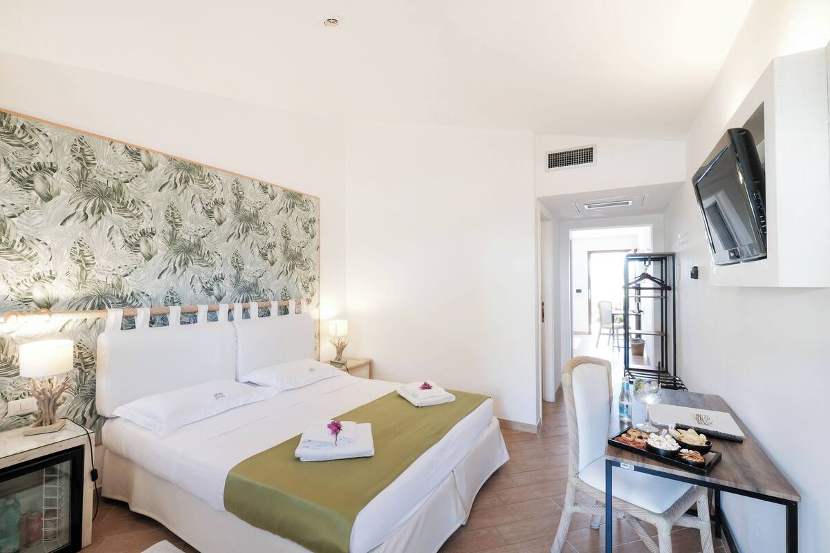 Quarto de hotel com cama, escrivaninha com laptop, televisão e porta que dá para uma área de refeições.