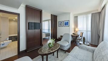 Quarto de hotel com sofá, poltrona e uma pequena mesa com um vaso de flores. Há uma televisão na parede e uma janela com cortinas.