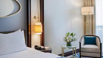 Quarto de hotel com cama, mesa de cabeceira com telefone, luminária, cadeira e um vaso com flores.