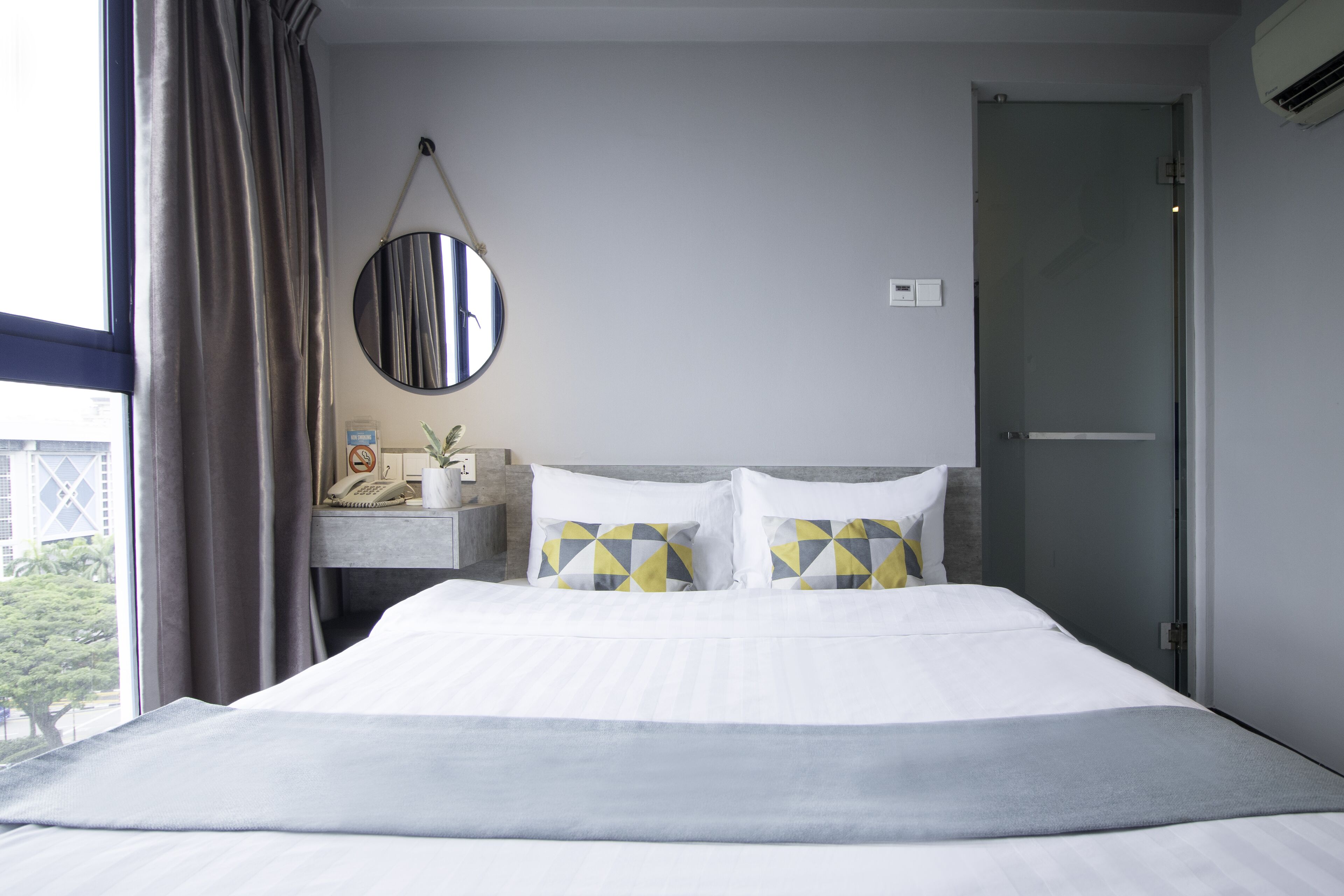 Quarto de hotel moderno com uma cama grande, mesa de cabeceira com abajur, espelho e vista para o exterior.