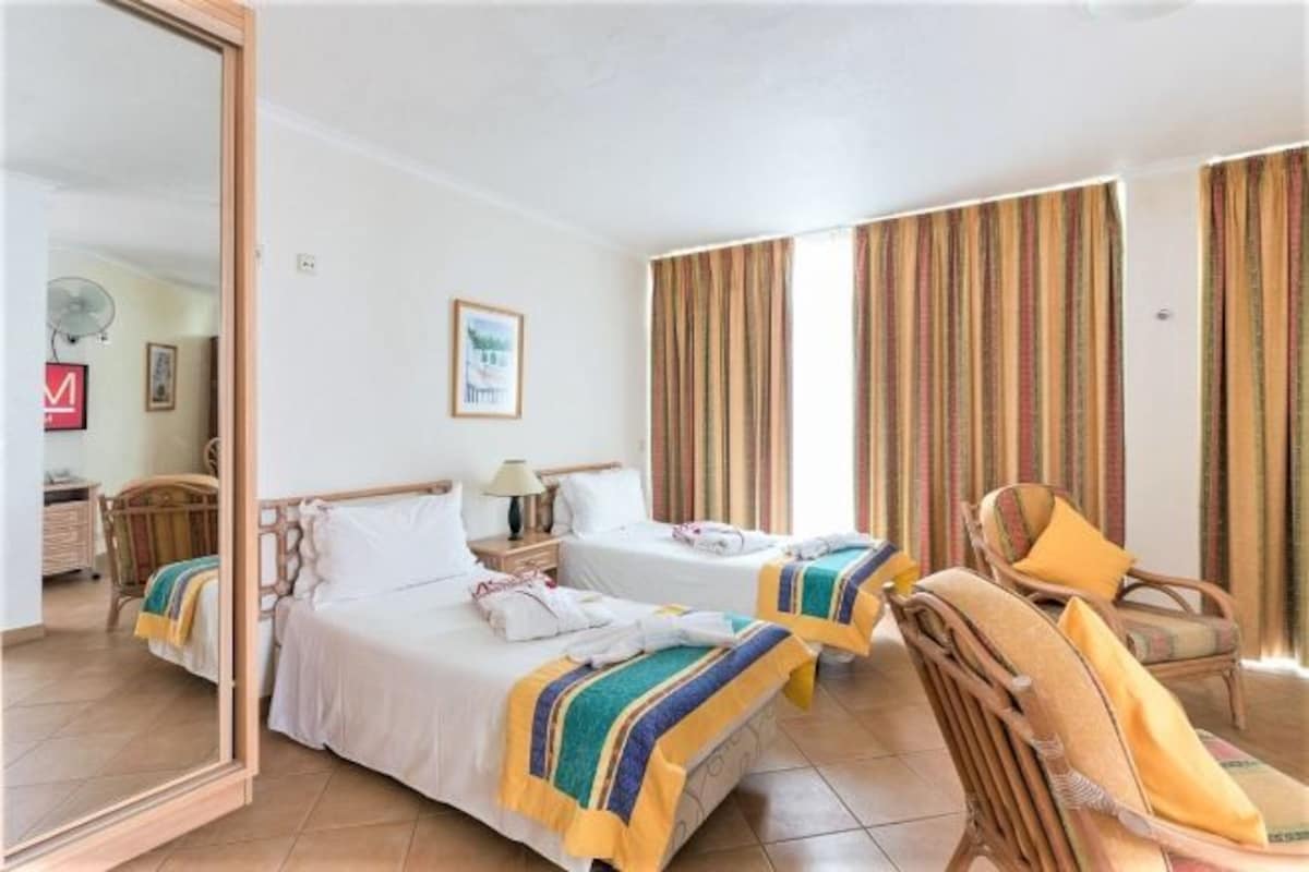 Quarto de hotel com duas camas, um espaço de estar com uma poltrona, um espelho e uma escrivaninha com abajur.