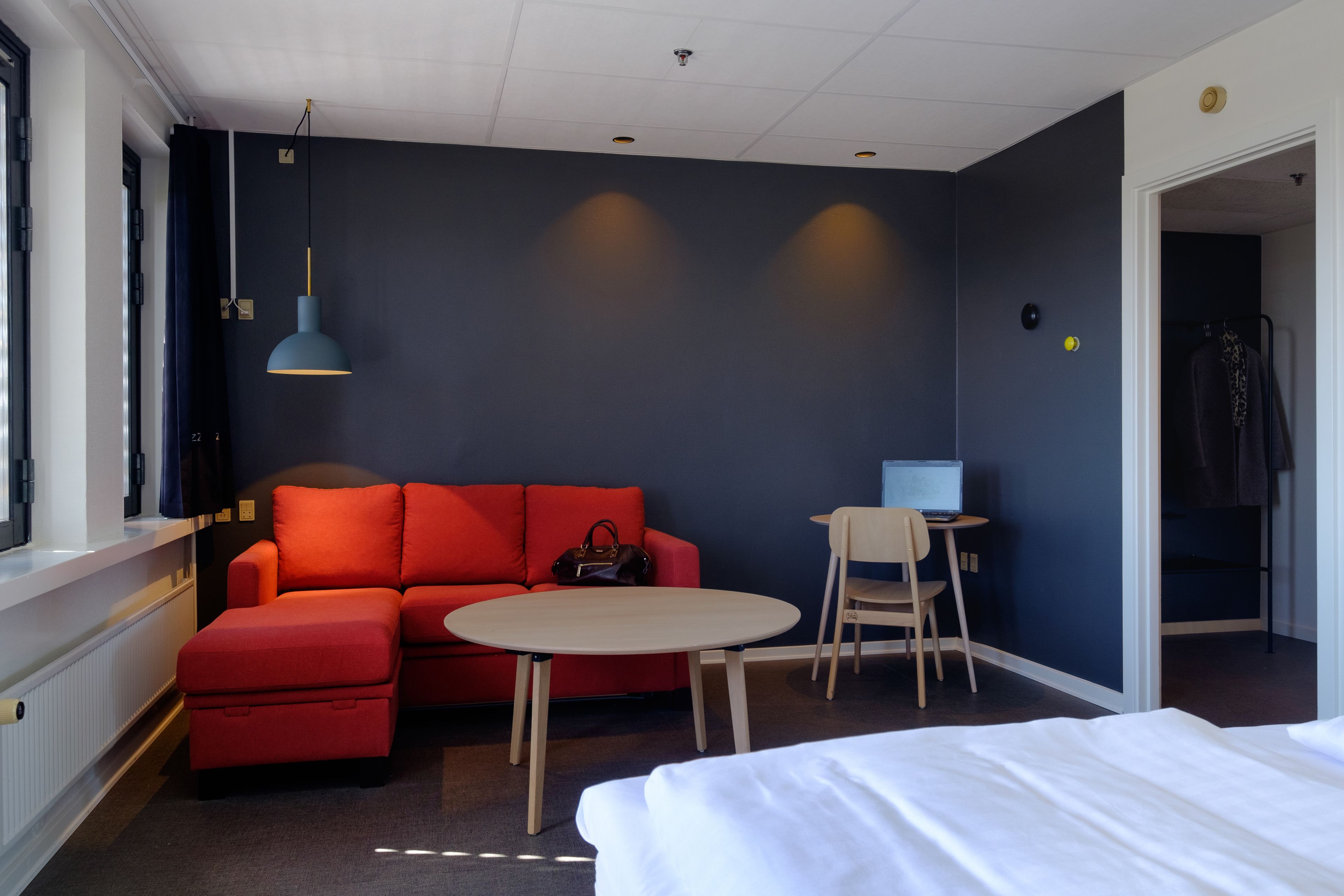 Quarto de hotel com sofá vermelho, uma mesinha, uma cama e uma escrivaninha com um laptop.