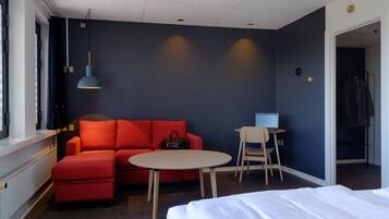 Quarto de hotel com sofá vermelho, uma mesinha, uma cama e uma escrivaninha com um laptop.