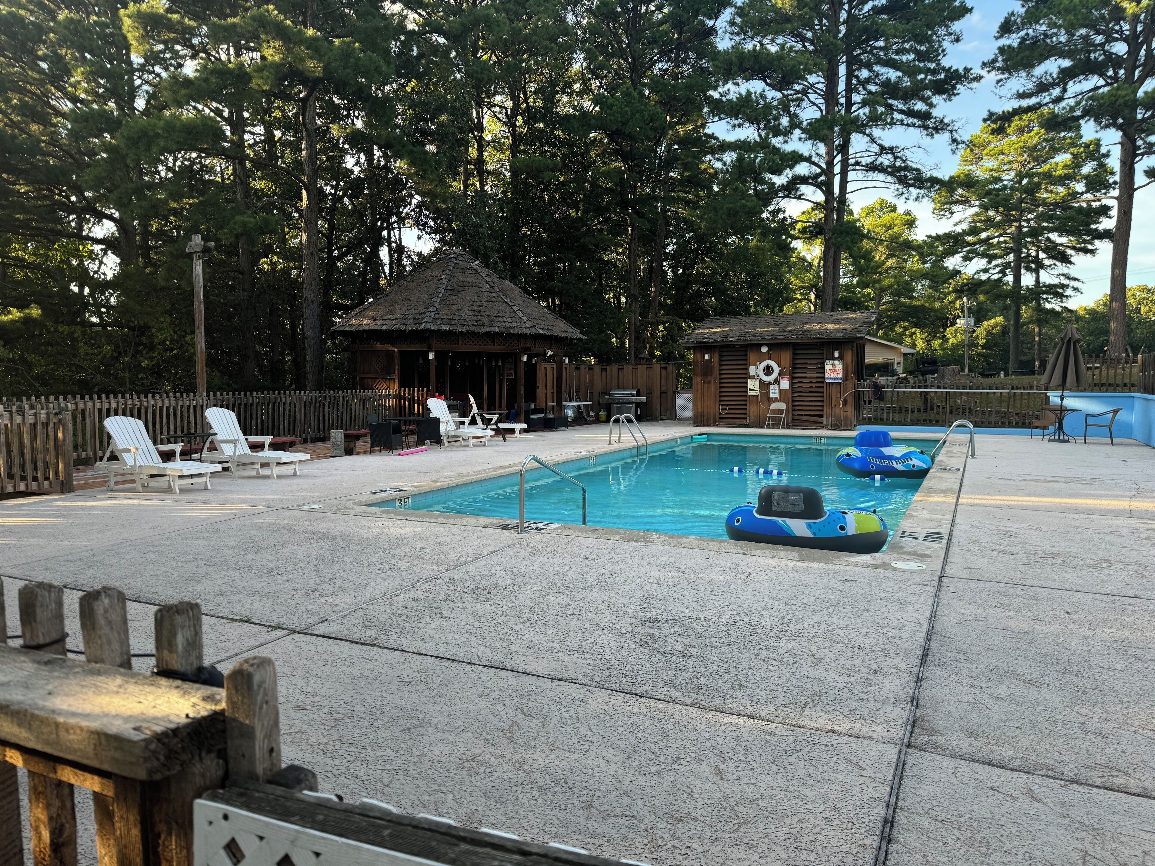 Piscina com espreguiçadeiras e brinquedos infláveis, cercada por um muro de madeira e árvores.