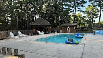 Piscina com espreguiçadeiras e brinquedos infláveis, cercada por um muro de madeira e árvores.