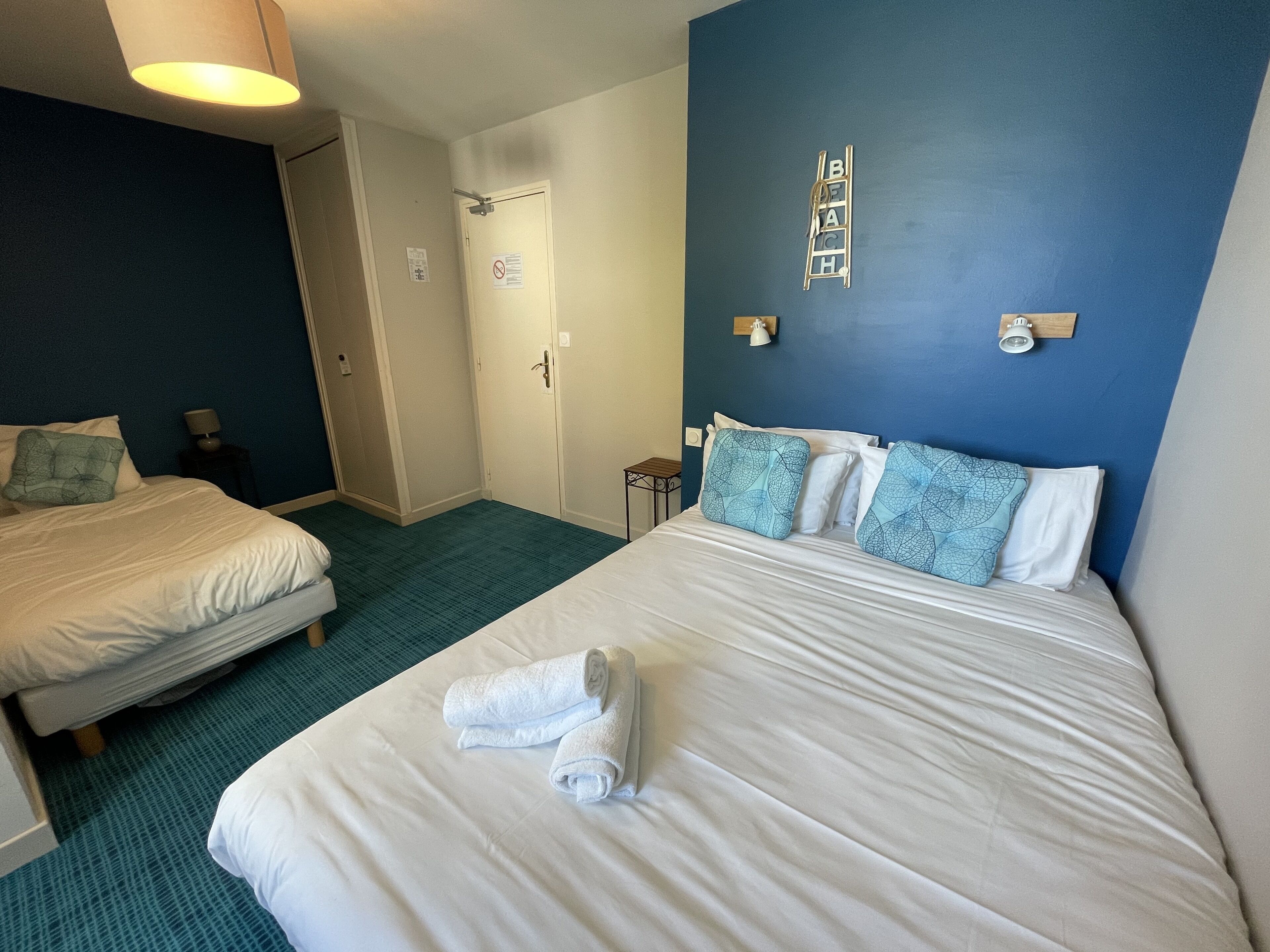 Quarto de hotel com duas camas, carpete verde-água, roupa de cama branca e uma parede de destaque azul.