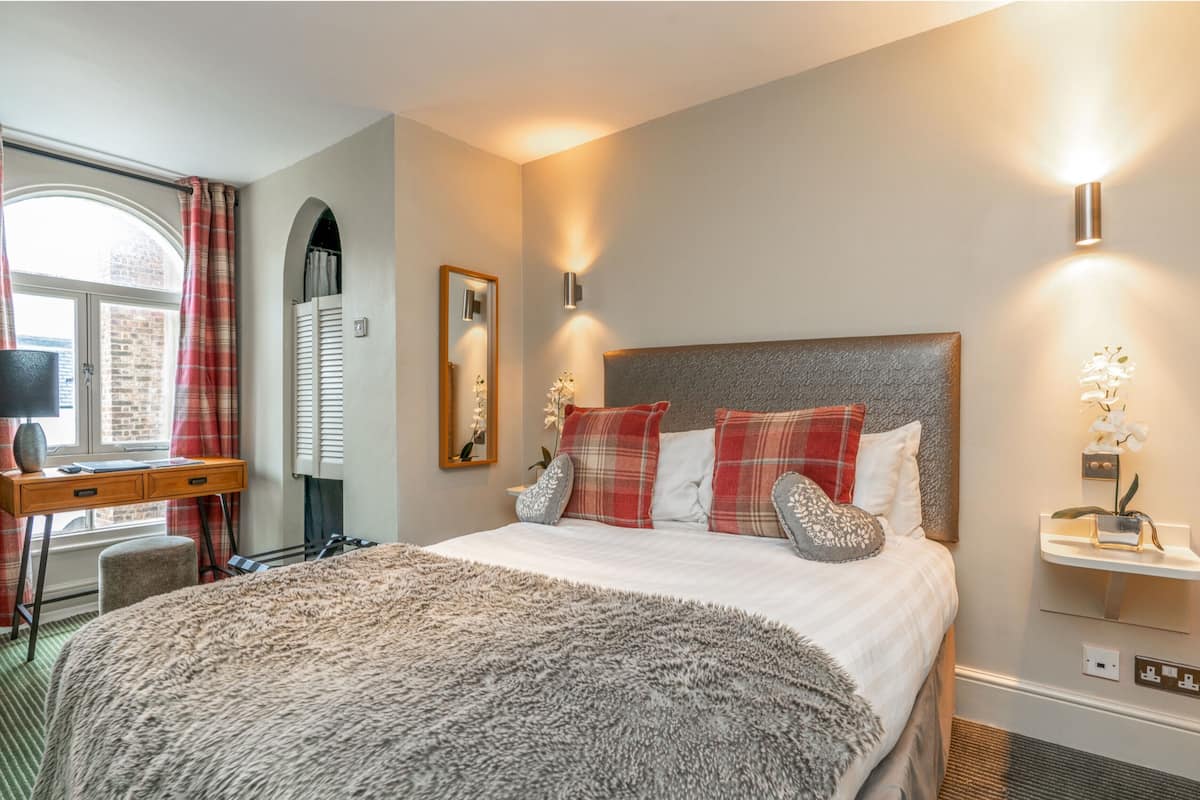 Quarto bem arrumado com cama, mesa de cabeceira, abajur e janela com cortinas.
