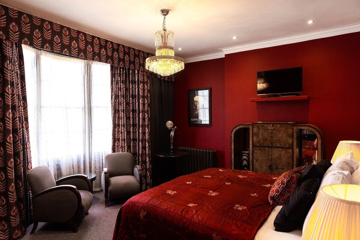 Quarto de hotel com uma cama grande, duas poltronas, um candelabro, uma televisão e um radiador.