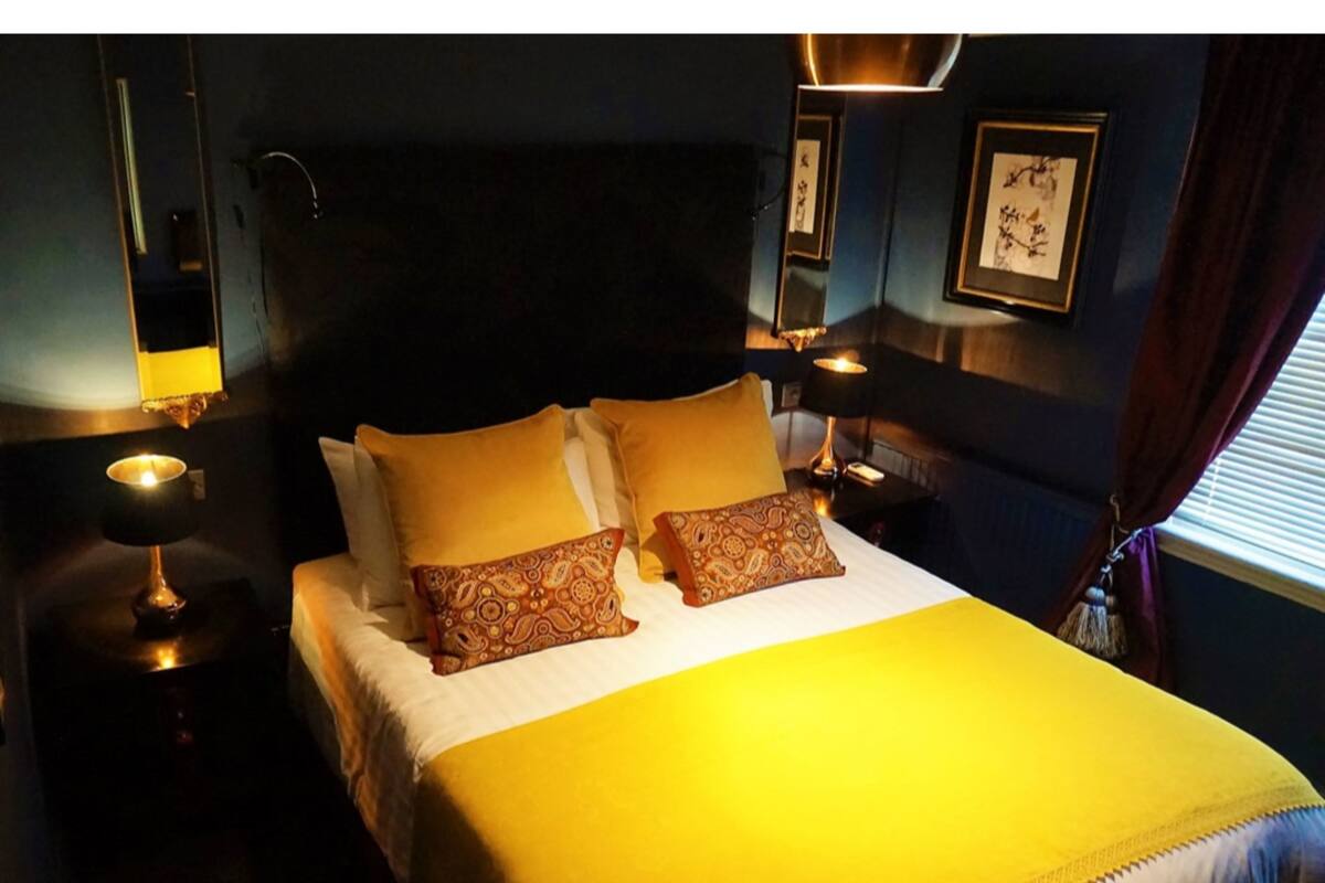 Quarto de hotel com cama, duas luminárias laterais, mesa de cabeceira e janela com cortinas.