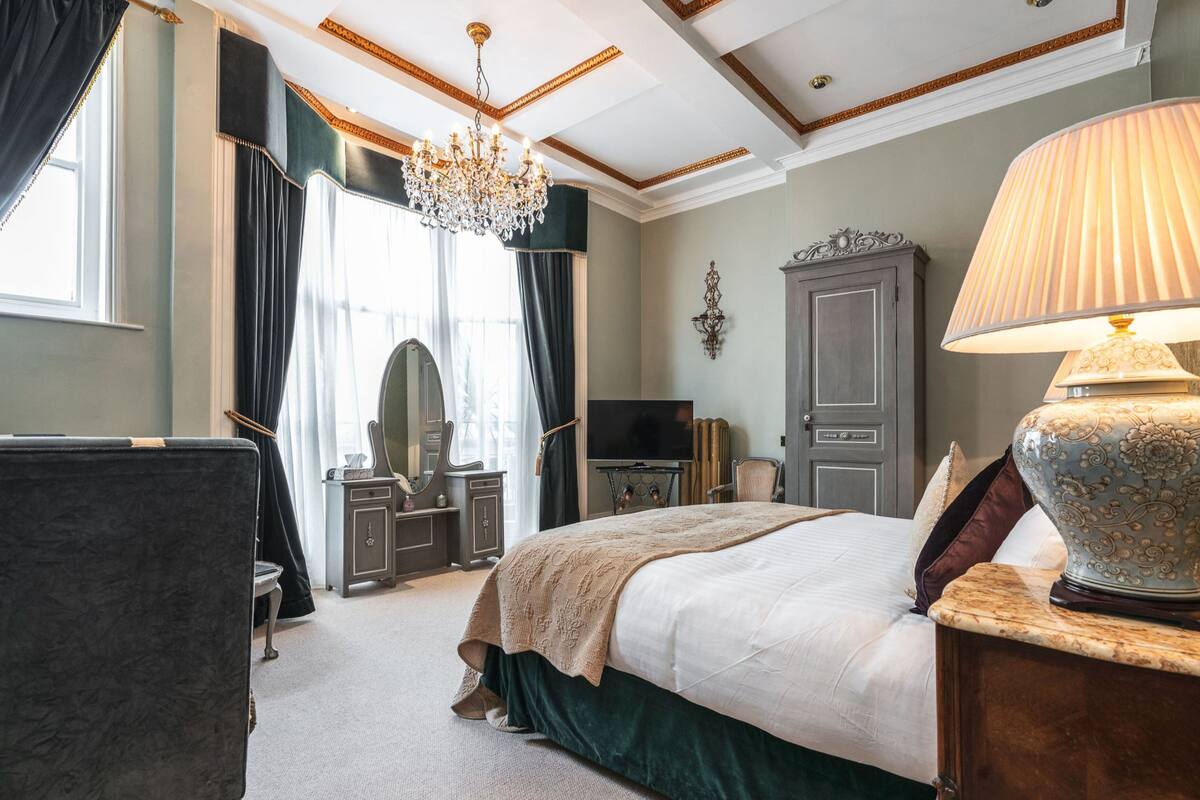 Quarto de hotel com cama, escrivaninha, cadeira, televisão, cômoda e um candelabro.