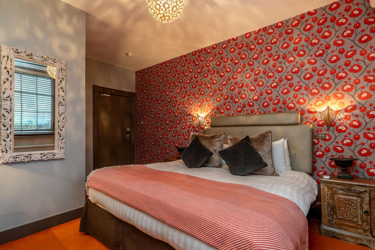 Quarto com cama grande, espelho decorativo, parede estampada e mesa de cabeceira com abajur.