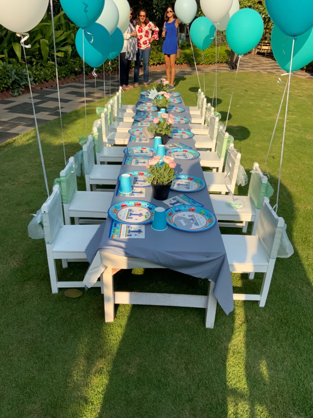 Mesa comprida montada ao ar livre, com cadeiras brancas, balões azuis e brancos e toalha de mesa com padrão azul e branco.