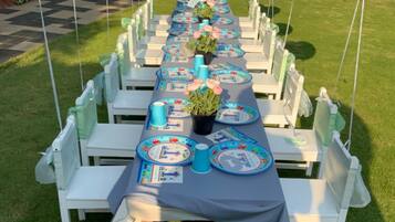 Mesa comprida montada ao ar livre, com cadeiras brancas, balões azuis e brancos e toalha de mesa com padrão azul e branco.