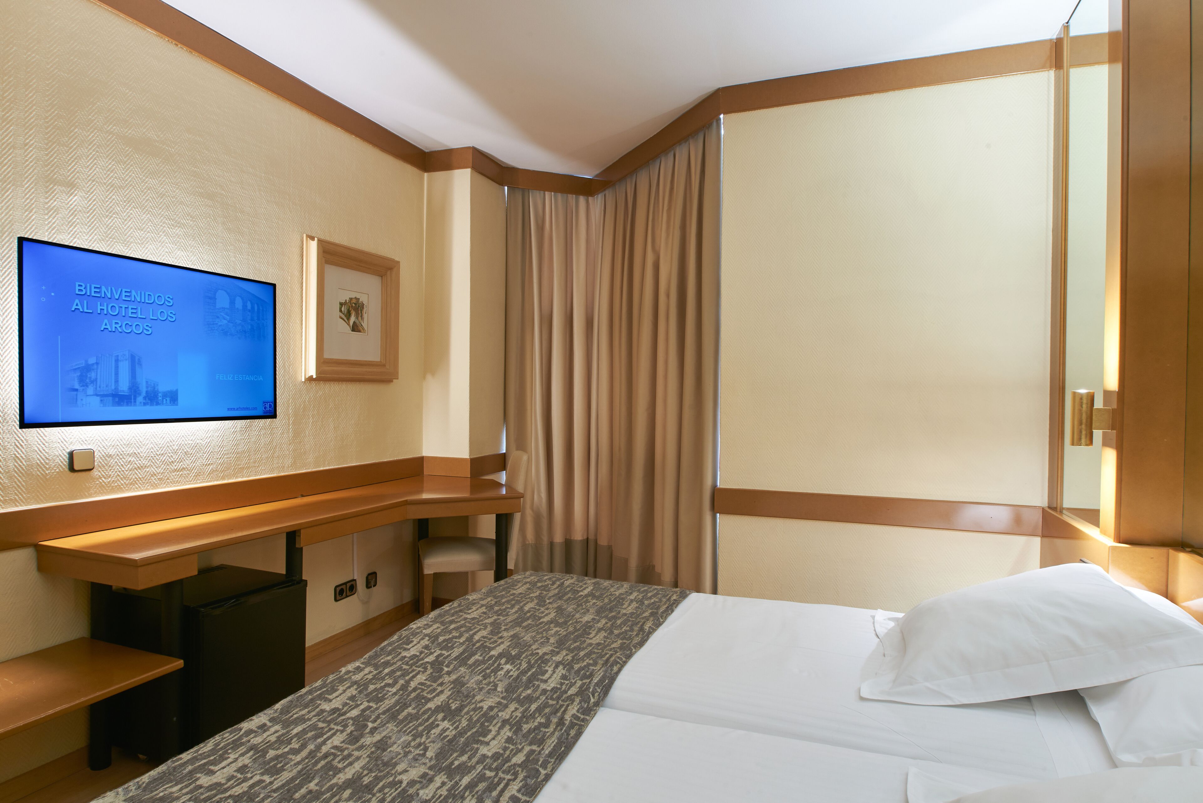 Quarto de hotel com uma cama, uma escrivaninha com cadeira, uma televisão exibindo uma mensagem de boas-vindas, um quadro na parede e uma cortina.