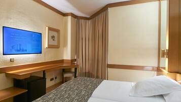 Quarto de hotel com uma cama, uma escrivaninha com cadeira, uma televisão exibindo uma mensagem de boas-vindas, um quadro na parede e uma cortina.