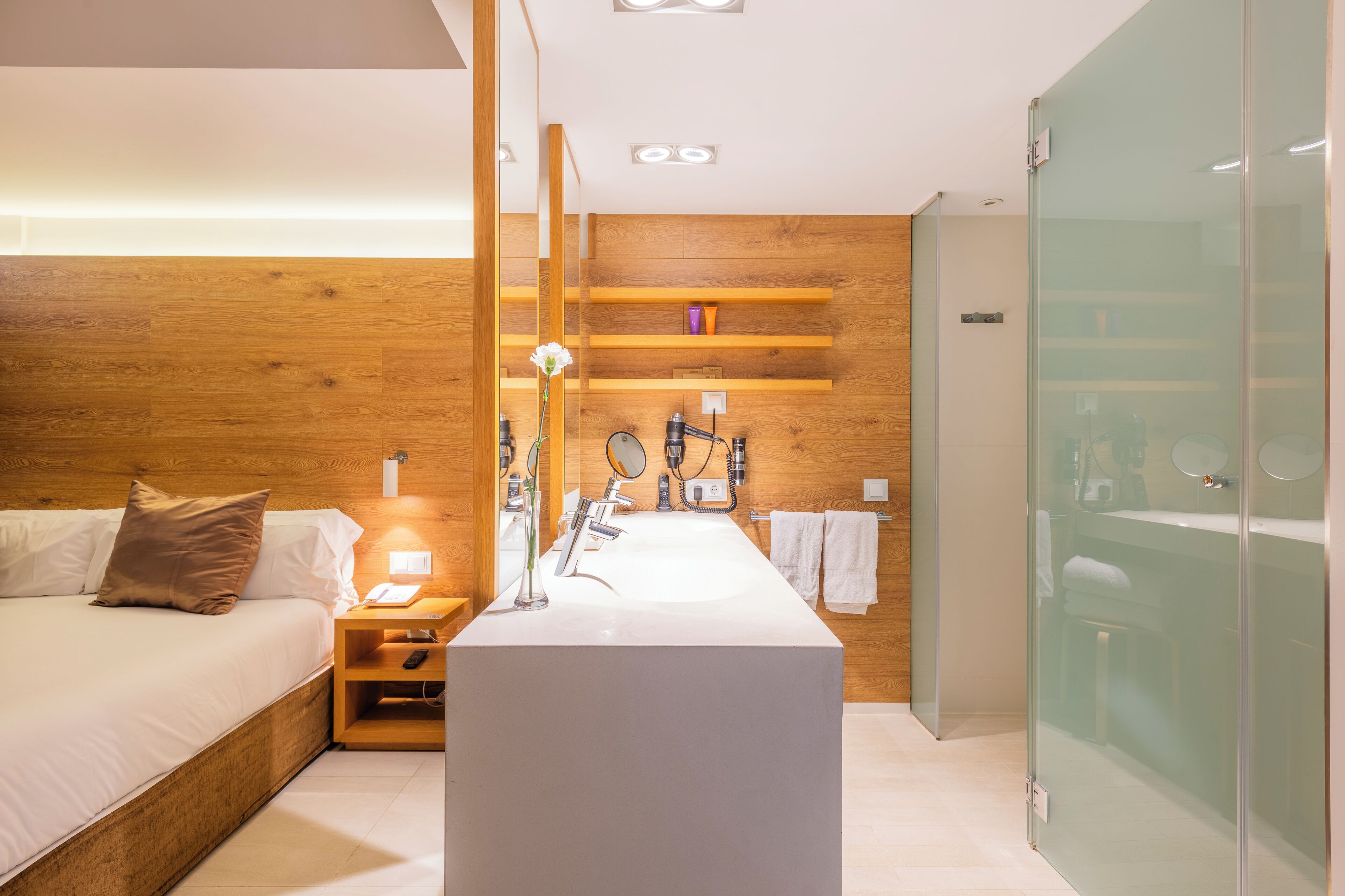 Quarto de hotel moderno com cabeceira de madeira, uma pia grande e branca, e box de chuveiro de vidro.