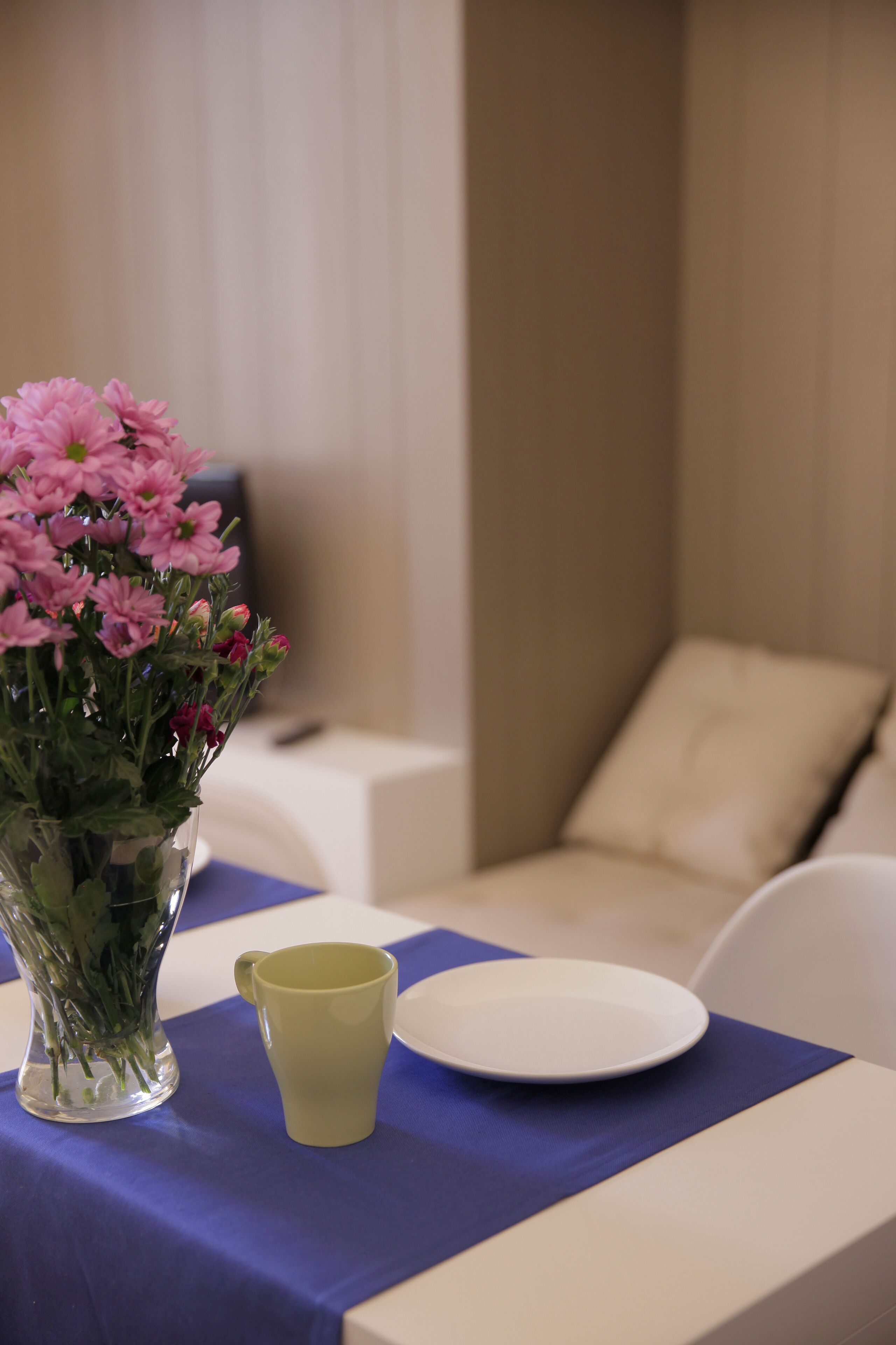 Uma mesa com um vaso de flores cor-de-rosa, uma caneca verde e um prato branco sobre uma toalha de mesa azul.