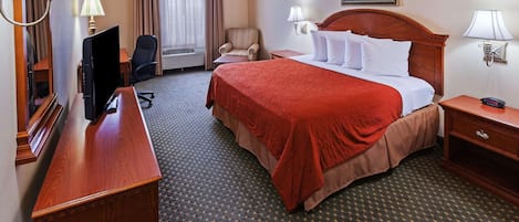 Quarto de hotel com uma cama grande, uma mesa com computador, uma cadeira, abajur e janela com cortinas.