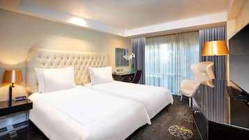 Quarto de hotel com uma cama grande, uma escrivaninha com cadeira, uma televisão e uma janela com cortinas.
