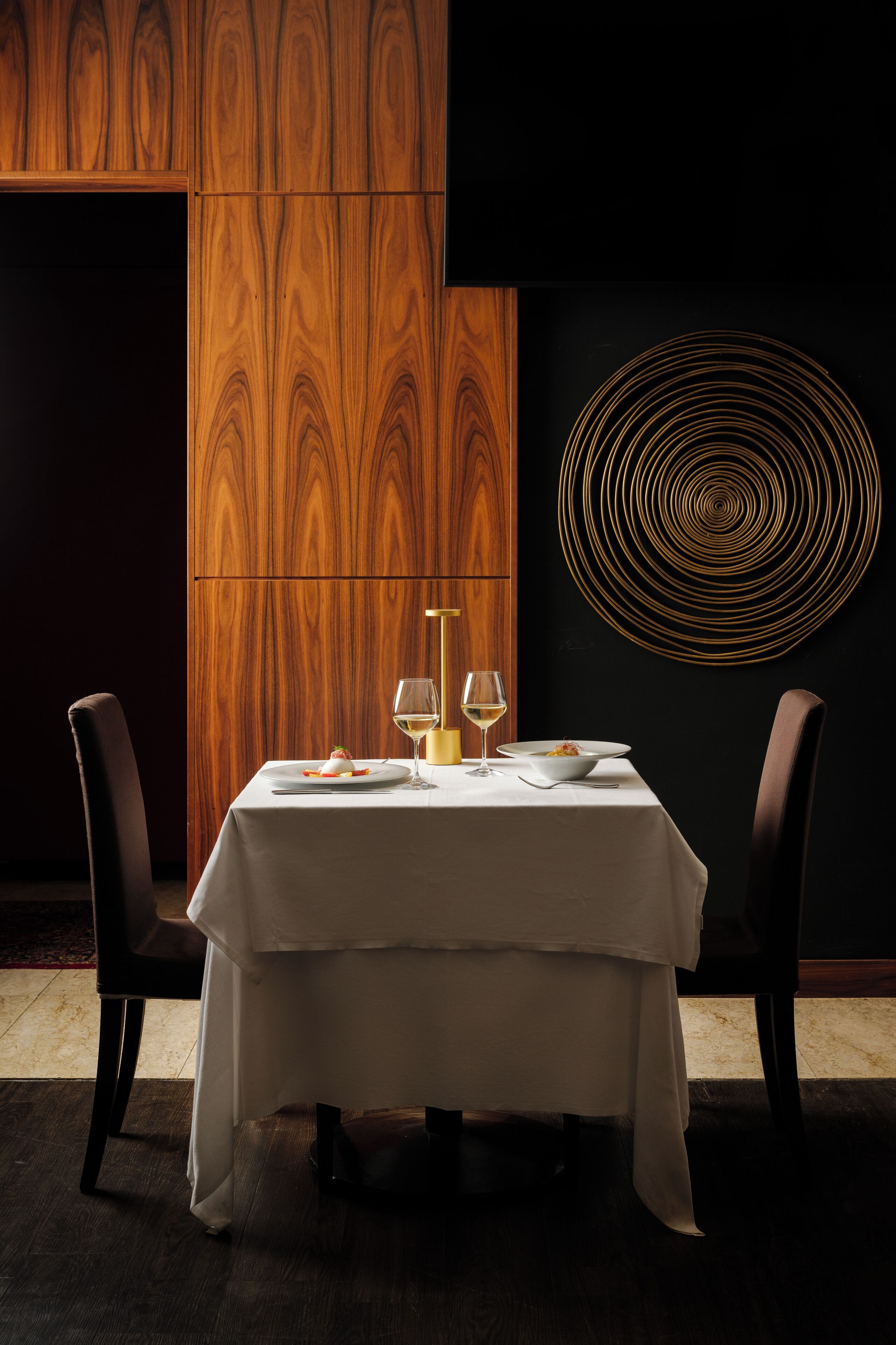 Uma área de refeições com uma mesa posta para dois, com toalha de mesa branca, duas cadeiras, uma taça de vinho e um prato de comida.