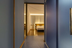 Corredor de hotel que leva a um quarto com uma cama, mesa de cabeceira e abajur.