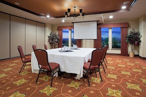 Uma sala de conferências com uma mesa branca, cadeiras vermelhas, uma tela de projetor e vista para a cidade ao entardecer.
