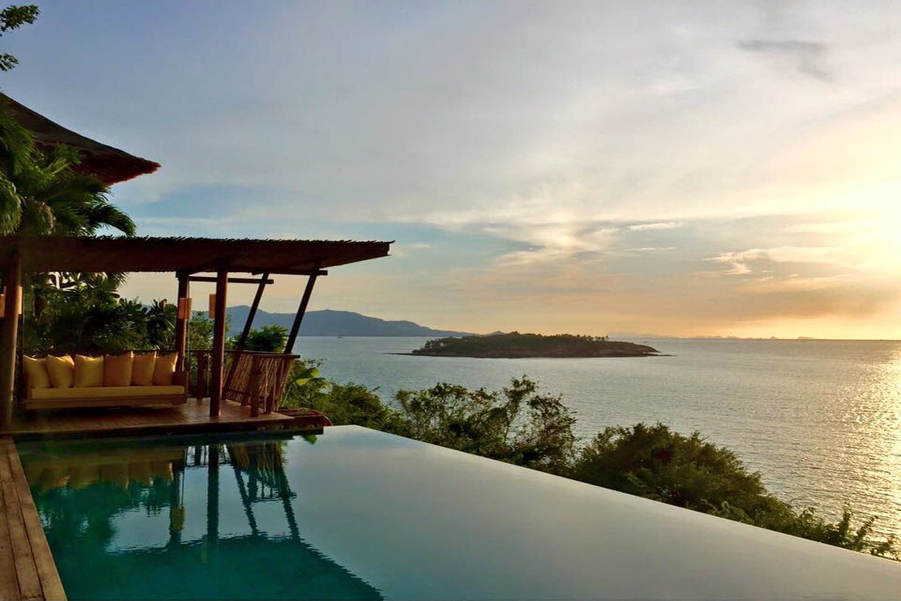 Piscina infinita com deck de madeira e área de estar com vista para um corpo d'água e terras distantes.