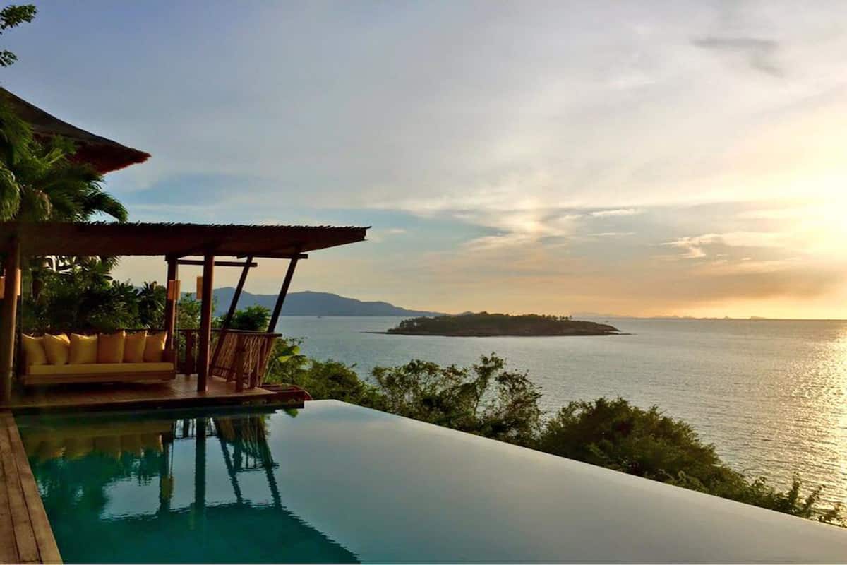 Piscina infinita com deck de madeira e área de estar com vista para um corpo d'água e terras distantes.