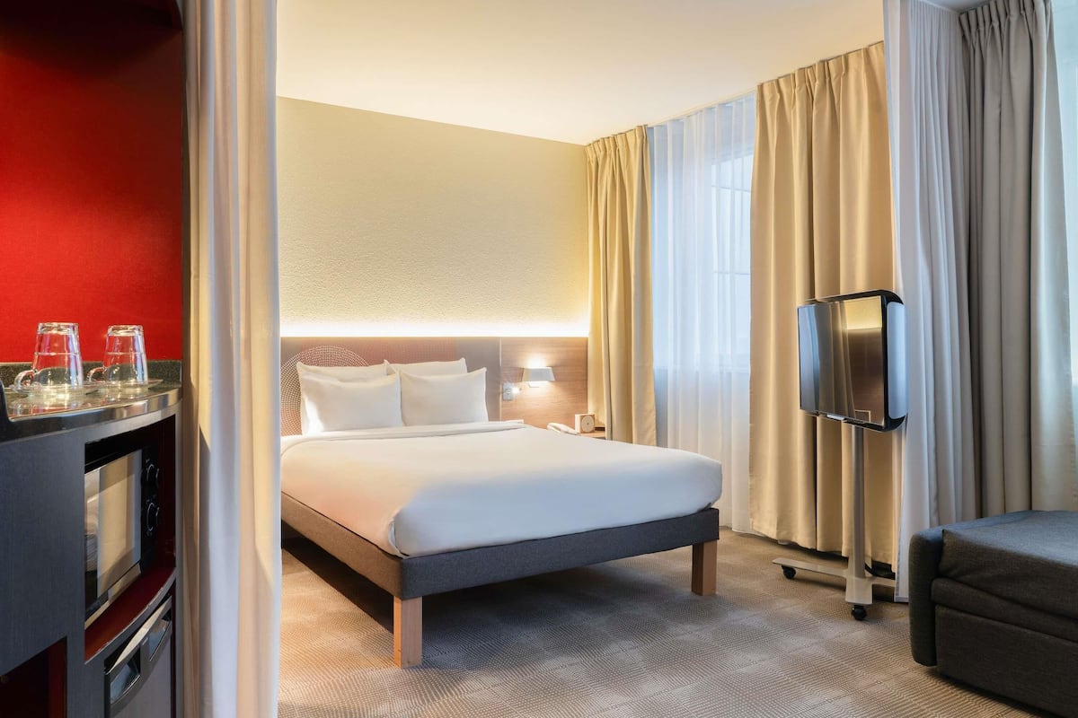 Quarto de hotel com cama, televisão, escrivaninha com cafeteira e janela com cortinas.