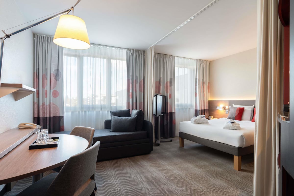 Quarto de hotel com cama, escrivaninha, cadeira, televisão e abajur.
