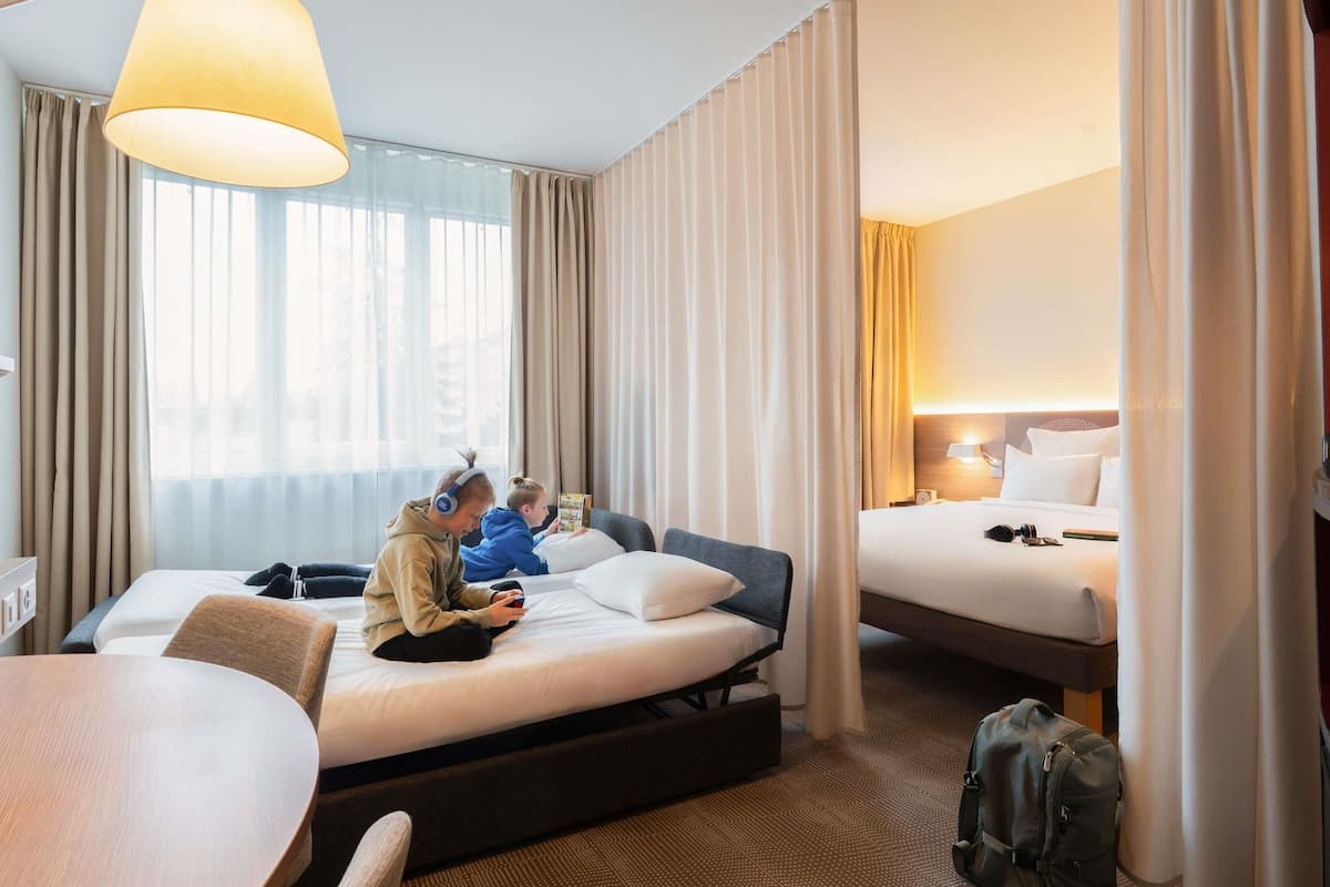 Quarto de hotel com duas camas, uma mesa de jantar e uma janela grande com cortinas.