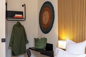 Quarto de hotel com uma cama, um casaco verde pendurado, uma pequena mesa com uma bolsa verde, uma prateleira na parede com livros e um adorno de parede.