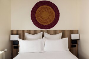 Quarto de hotel com uma cama, duas luminárias de cabeceira, uma obra de arte circular pendurada na parede e uma cabeceira de madeira.